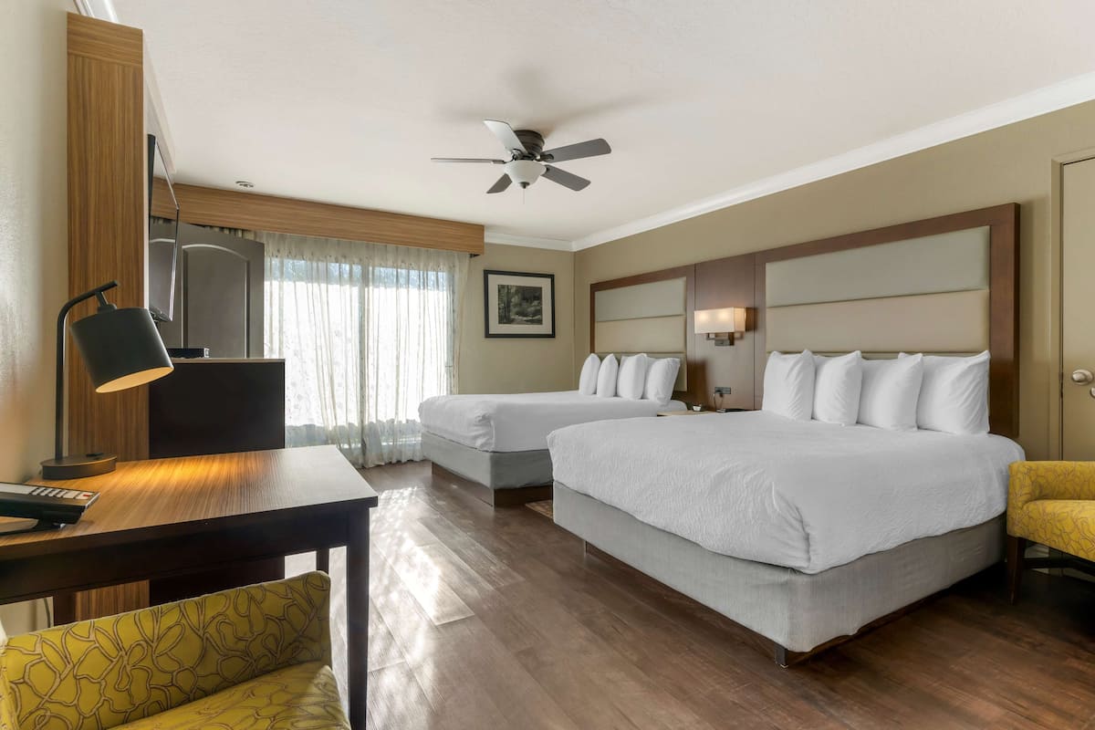 Quarto de hotel com duas camas, uma escrivaninha com lâmpada, uma cadeira, um ventilador de teto e uma janela com cortinas.