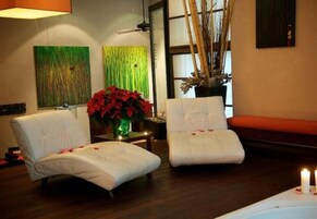 Um quarto com duas cadeiras brancas acolchoadas, uma pequena mesa com um vaso de flores vermelhas e uma grande janela com detalhes em bambu.