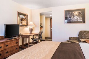 Quarto de hotel com cama, escrivaninha com luminária, televisão e uma cadeira.