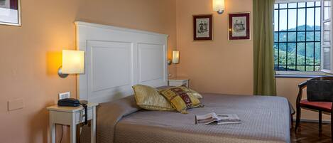 Uma cama bem arrumada com colcha, uma mesa de cabeceira com luminária e uma janela com vista para o exterior.