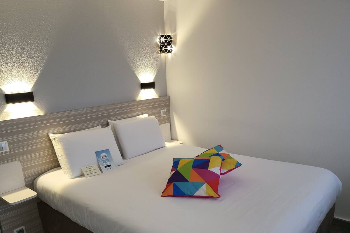 Cama bem arrumada com lençóis brancos e duas almofadas geométricas coloridas, mesa de cabeceira com abajur e luminária fixada na parede.