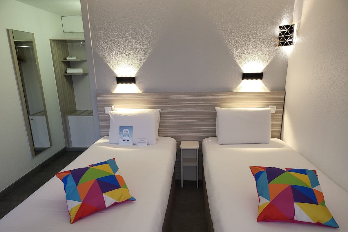 Quarto de hotel com duas camas, cada uma com almofada geométrica colorida, uma mesa de cabeceira, um espelho e luminárias fixadas na parede.