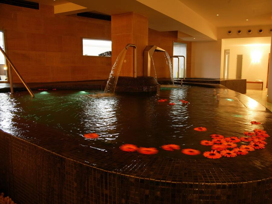 Uma grande piscina interior com uma fonte central, rodeada por um chão em mosaico.