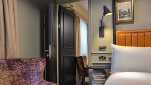 Quarto de hotel com cama, cadeira, escrivaninha com abajur e estante de livros.