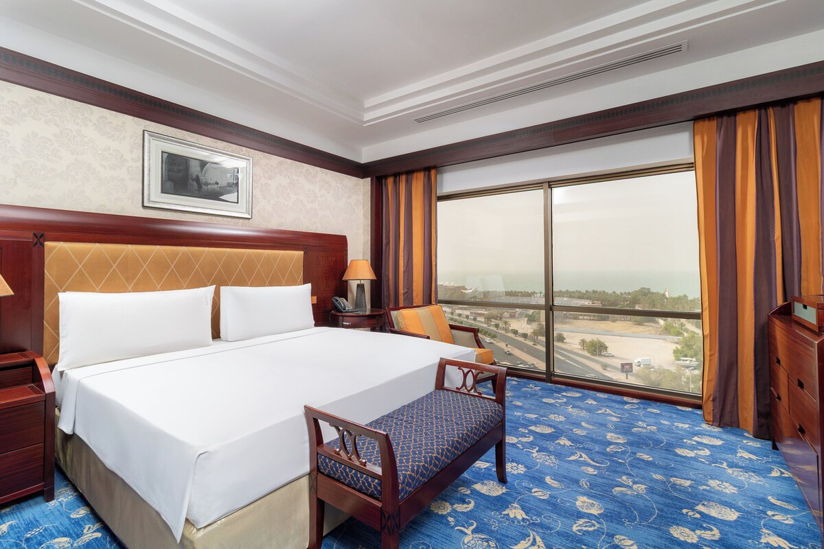 Quarto de hotel com uma cama grande, cabeceira de madeira, janela com cortinas, mesa de cabeceira, abajur e vista para a cidade.