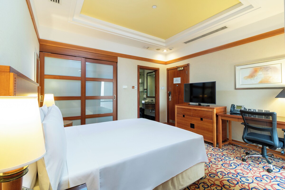 Quarto de hotel com cama, escrivaninha, cadeira, televisão e um tapete estampado.