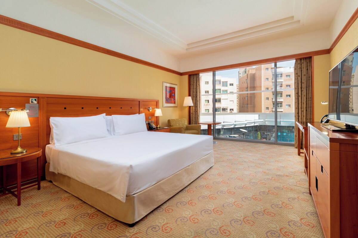 Quarto de hotel com uma cama grande, duas luminárias de cabeceira, uma escrivaninha com luminária, uma cadeira, uma televisão e vista da cidade através de amplas janelas.