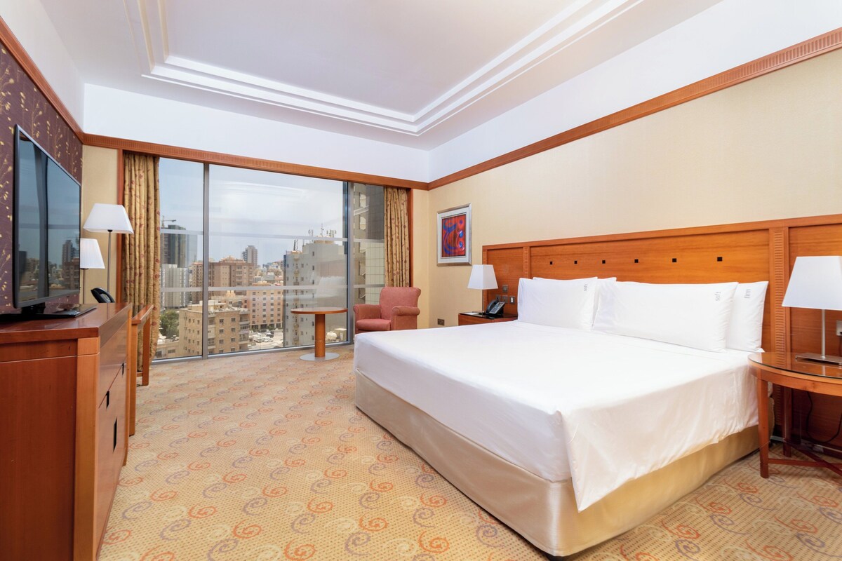 Quarto de hotel com uma cama grande, uma escrivaninha, uma cadeira, uma TV e vista da cidade por grandes janelas.