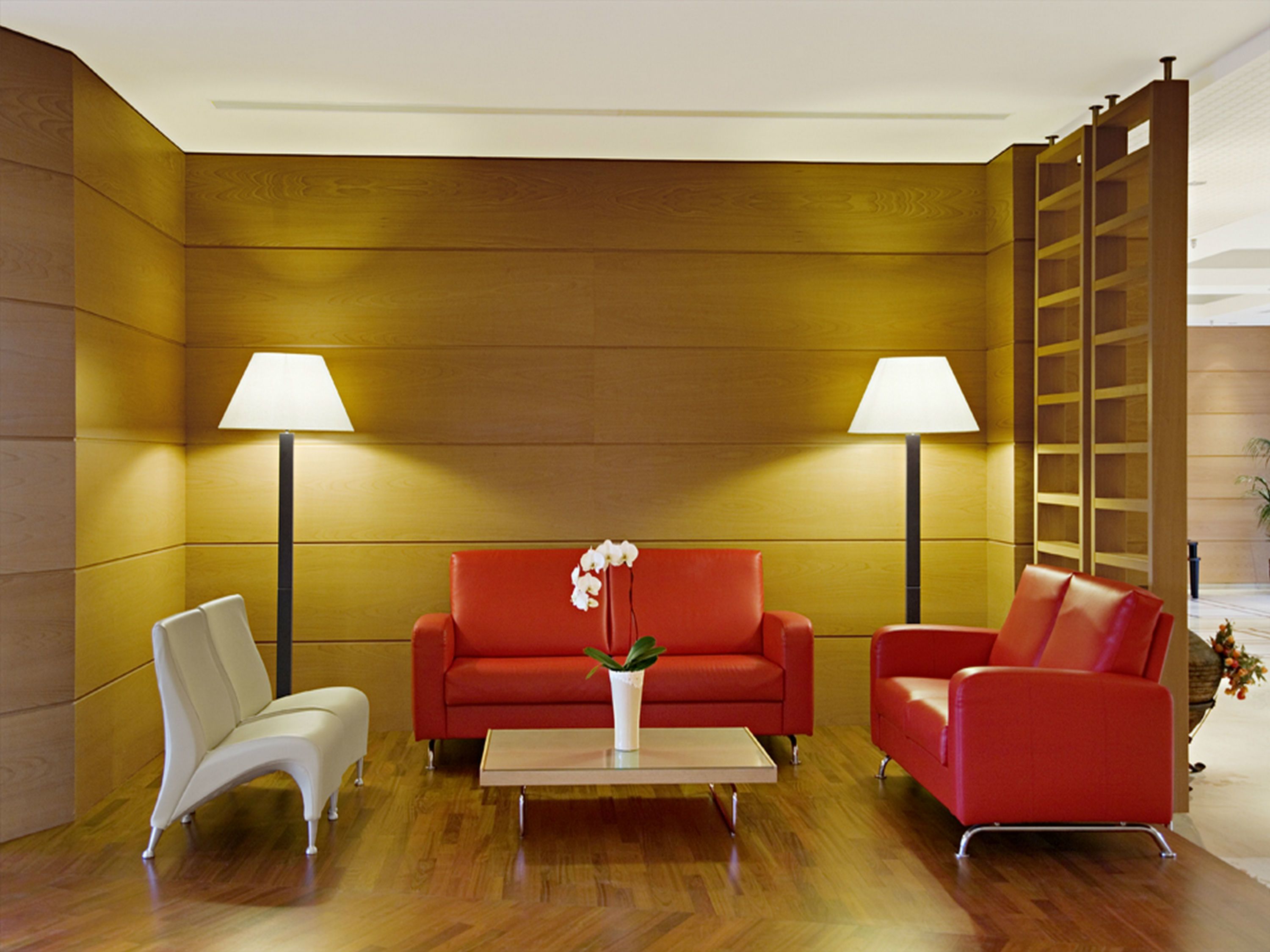 Uma sala de estar moderna com um sofá vermelho, uma poltrona branca e uma mesa de centro. Há duas luminárias de chão e uma estante de livros ao fundo.