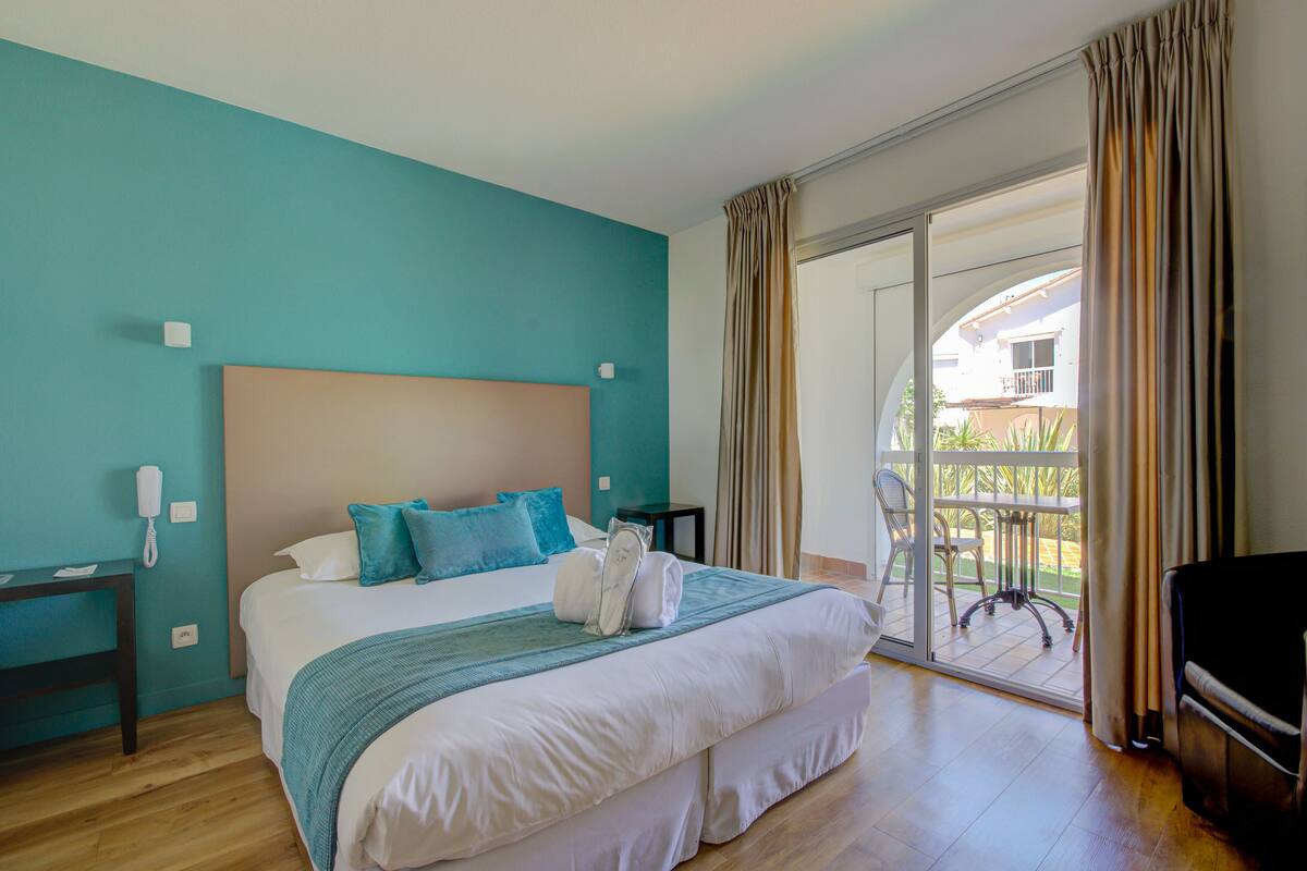 Quarto de hotel com uma cama grande, uma mesa de cabeceira com telefone, uma cadeira perto da janela e uma varanda com vista para o exterior.