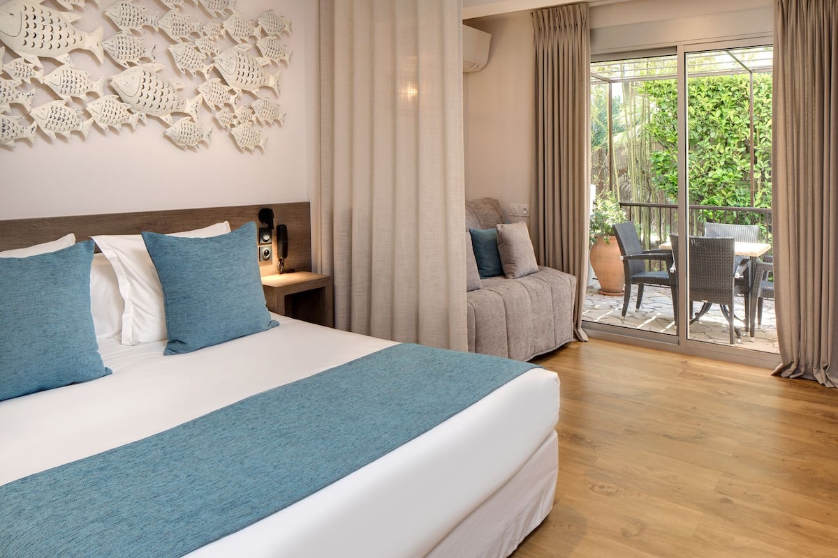 Quarto de hotel com uma cama grande, mesa de cabeceira, escrivaninha e vista para um pátio externo com cadeiras e mesa.