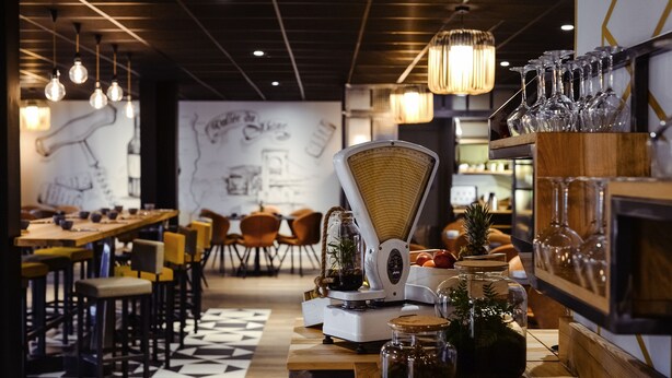 Interior moderno de restaurante com uma longa mesa de madeira, cadeiras, luminárias pendentes e uma parede com desenhos artísticos.