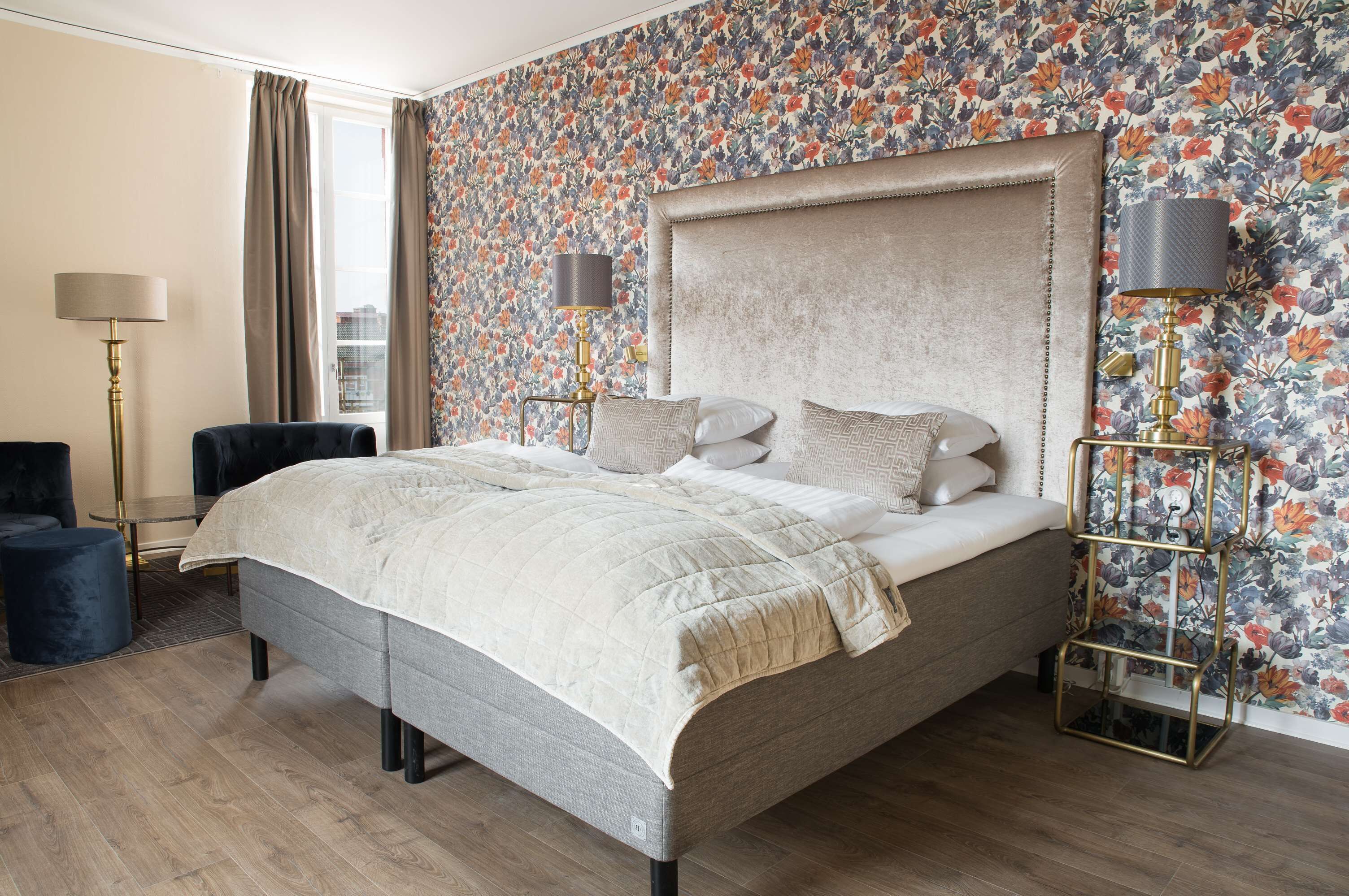 Quarto com uma cama grande, duas mesas de cabeceira com luminárias, uma cadeira e papel de parede floral.