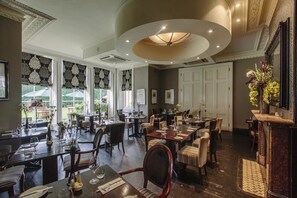 Uma ampla sala de jantar com várias mesas e cadeiras, grandes janelas com cortinas estampadas e um lustre central.