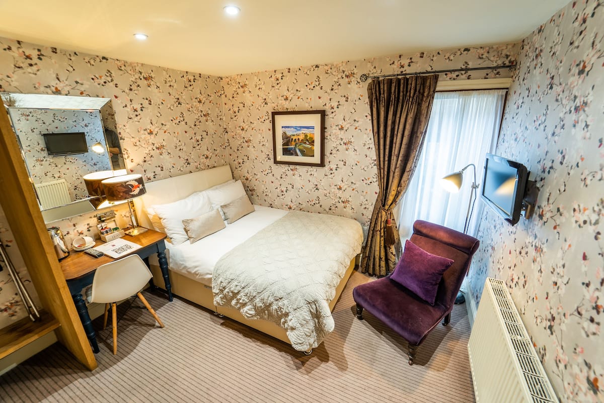 Quarto de hotel com cama, escrivaninha com cadeira, televisão e janela com cortinas.