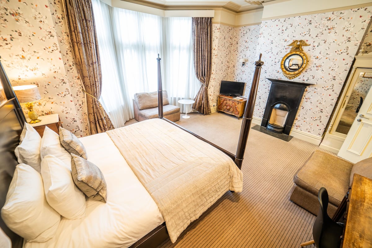 Quarto de hotel com cama, poltrona, televisão, lareira e espelho na parede.