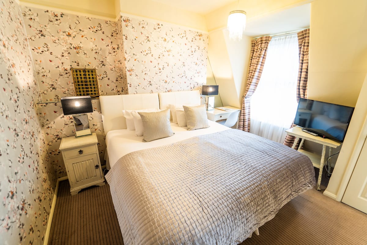 Quarto de hotel bem arrumado, com cama, mesas de cabeceira, televisão e janela com cortinas.