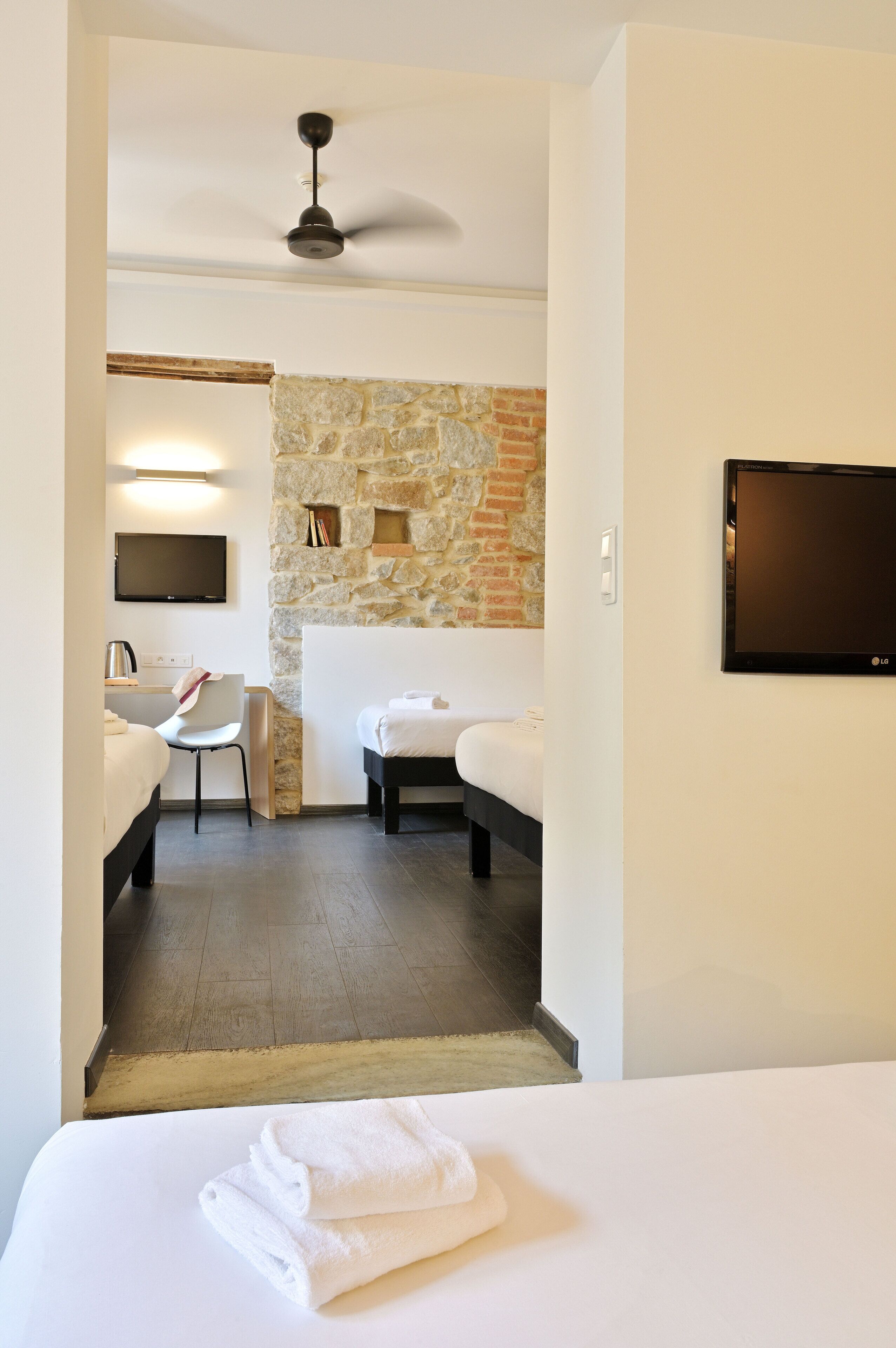 Quarto de hotel com parede de destaque em pedra, televisão de tela plana, cama com roupa de cama branca, uma cadeira e uma mesa.