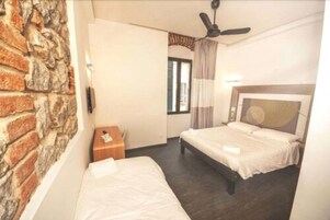 Quarto de hotel com parede de tijolos, ventilador de teto, cama, mesa de cabeceira e janela com cortinas.