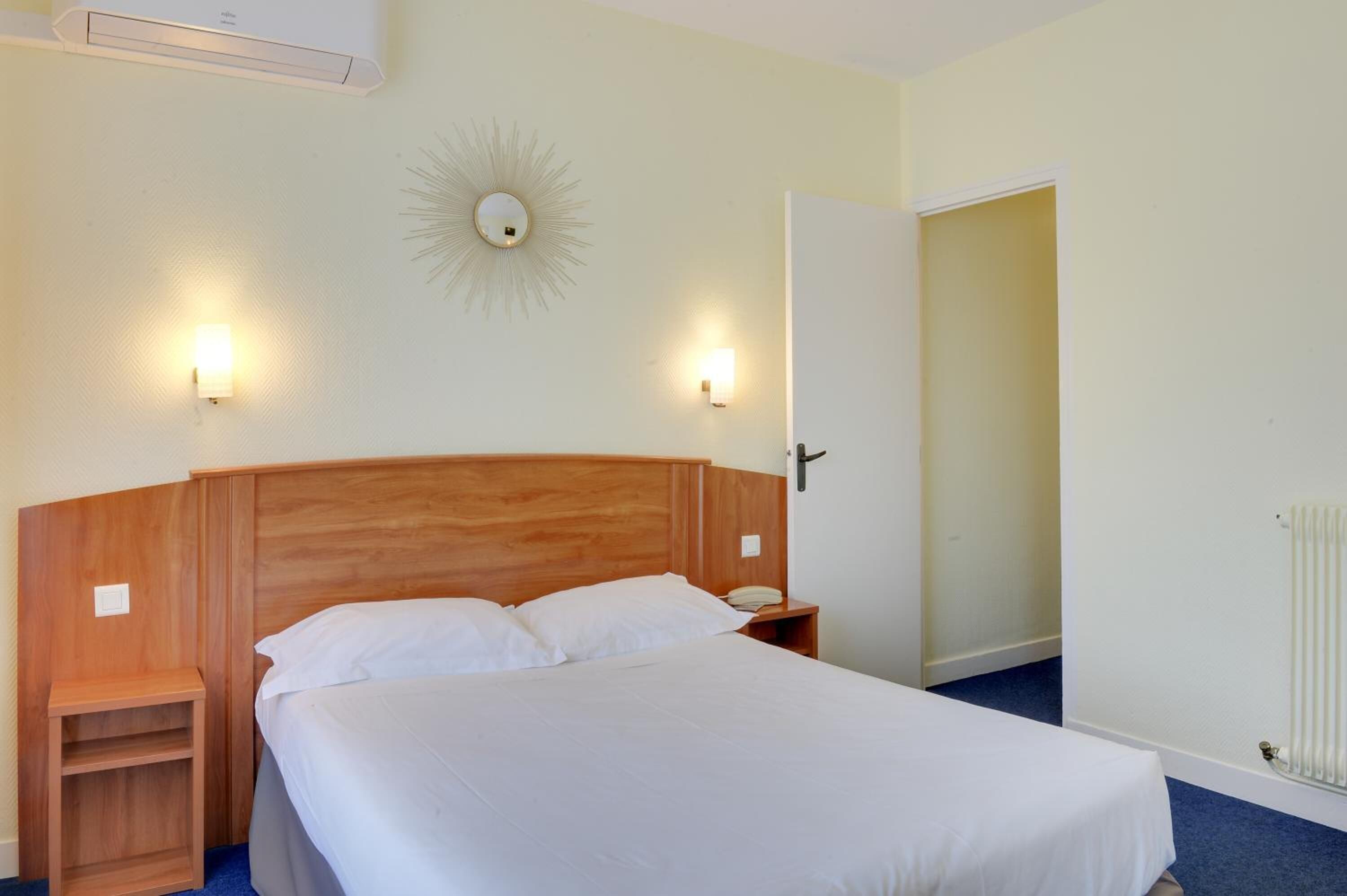 Quarto de hotel com cabeceira de madeira, uma cama de solteiro, duas mesas de cabeceira, um espelho fixado na parede e uma porta que dá para outro cômodo.
