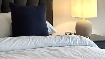 Uma cama bem arrumada com lençóis brancos e um travesseiro azul escuro, um abajur redondo com base texturizada e uma cabeceira estofada.