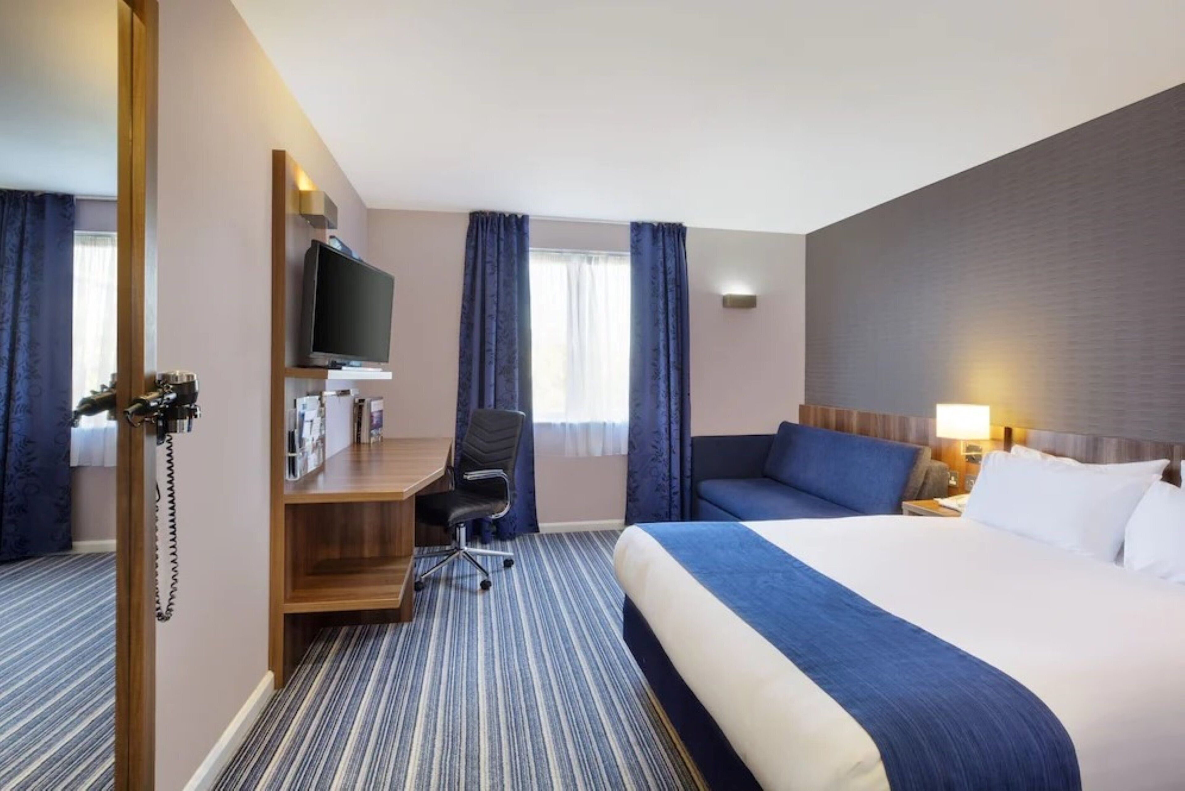 Quarto de hotel com cama, escrivaninha, cadeira, televisão e mesa de cabeceira com abajur.