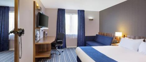 Quarto de hotel com cama, escrivaninha, cadeira, televisão e mesa de cabeceira com abajur.
