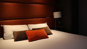 Cama bem arrumada com almofadas brancas e castanhas, uma cabeceira moderna e um abajur.