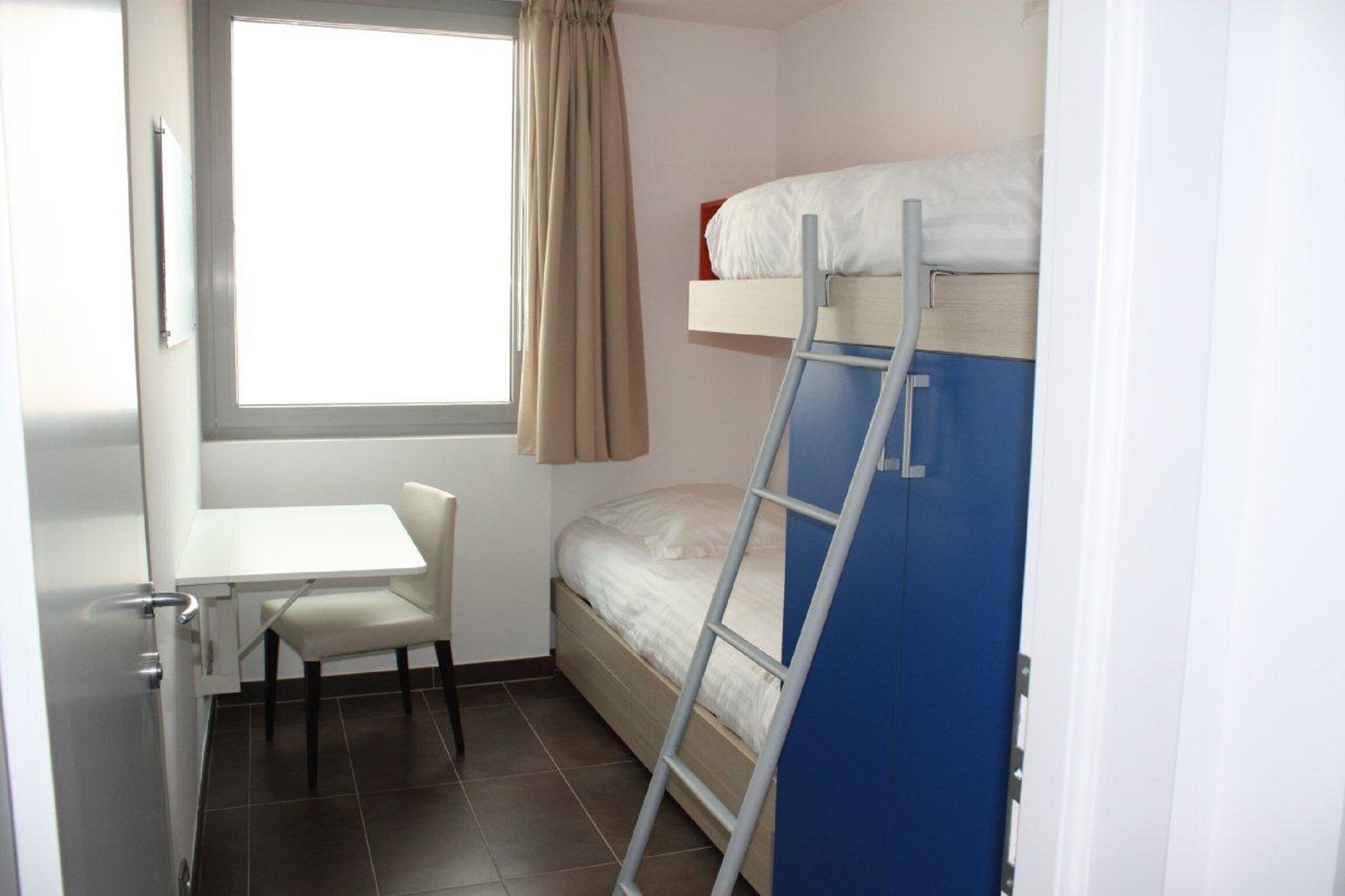 Quarto pequeno e compacto com uma cama de solteiro, mesa e cadeira.