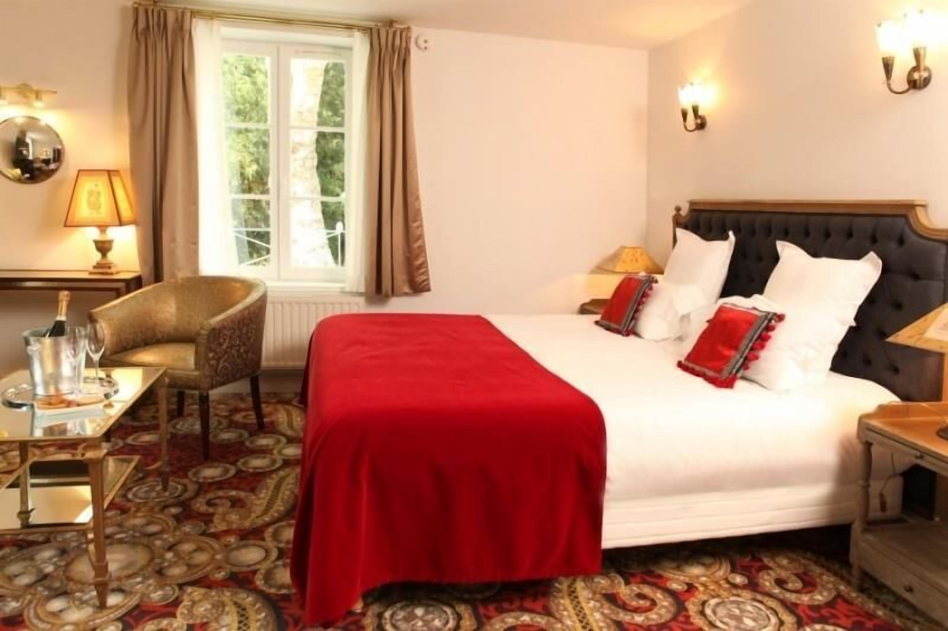 Quarto de hotel com uma cama grande, área de estar com uma poltrona, mesa com abajur e janela com cortinas.