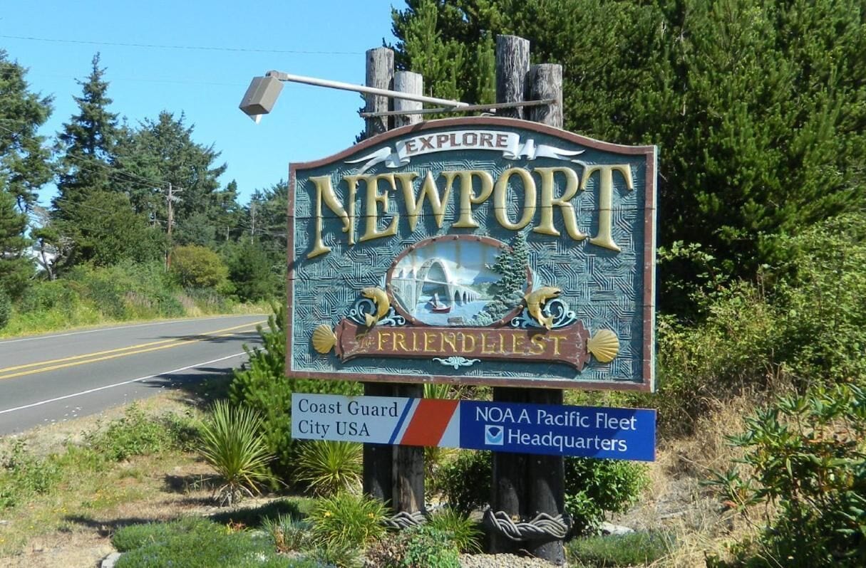 Placa indicando Newport, identificada como 'The Friendlyest', com vista panorâmica do oceano e das montanhas.