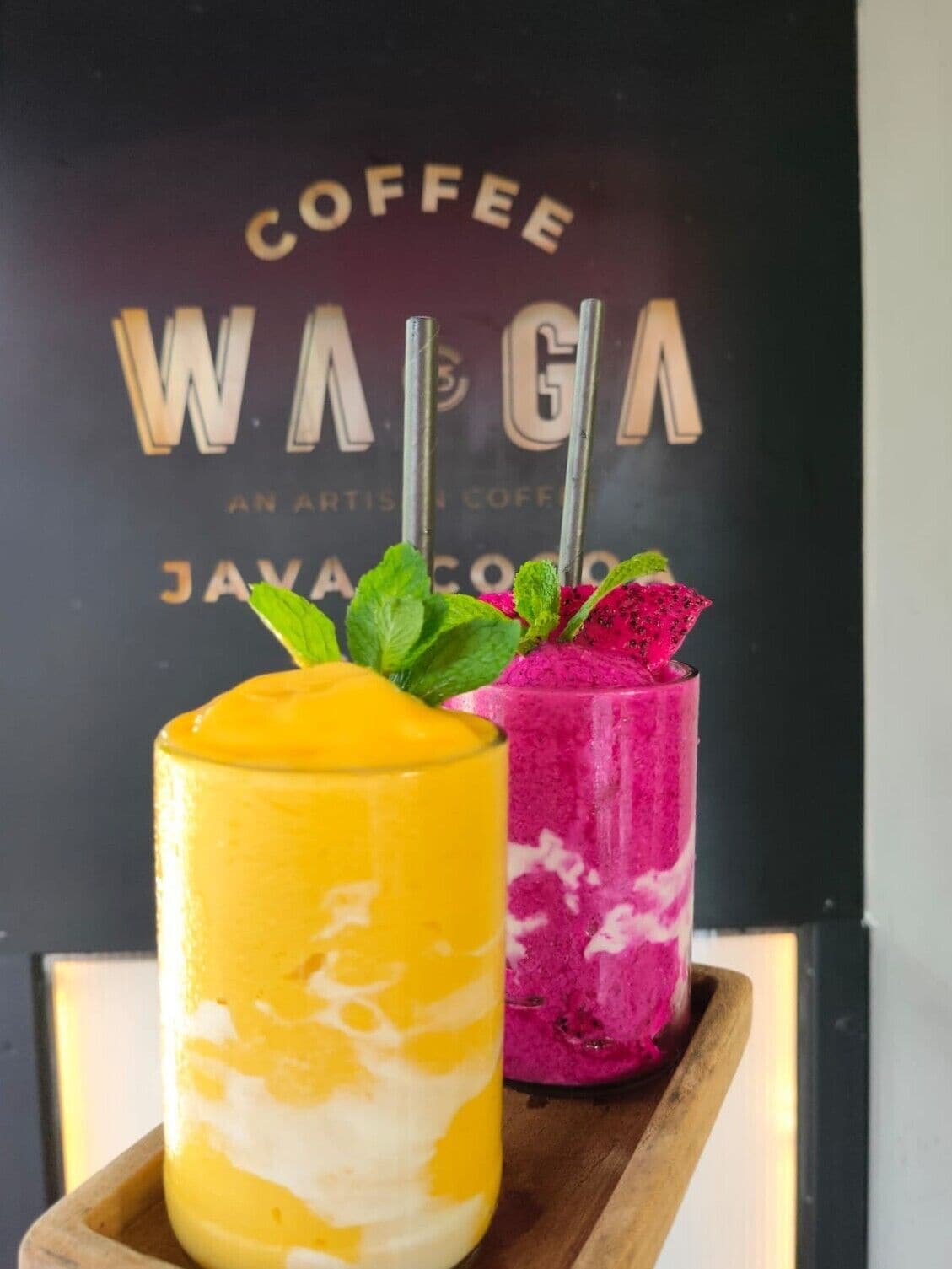 Duas bebidas geladas com canudos e folhas de hortelã sobre uma bandeja de madeira, com uma placa ao fundo que diz 'Coffee WaGA'.