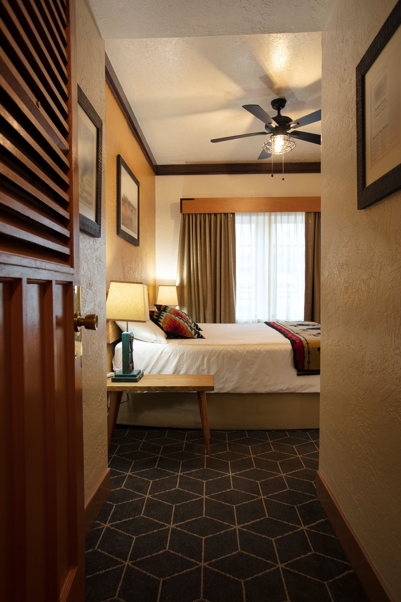 Quarto de hotel com cama, escrivaninha com luminária, ventoinha no teto e janela com cortinas.