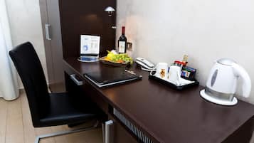 Mesa de quarto de hotel com um laptop, uma garrafa de vinho, uma cesta de frutas, um telefone, uma bandeja com copos e lanches, e uma chaleira.