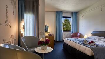 Quarto de hotel com uma cama, duas cadeiras, uma mesinha com um vaso de flores, uma televisão e uma janela com cortinas.