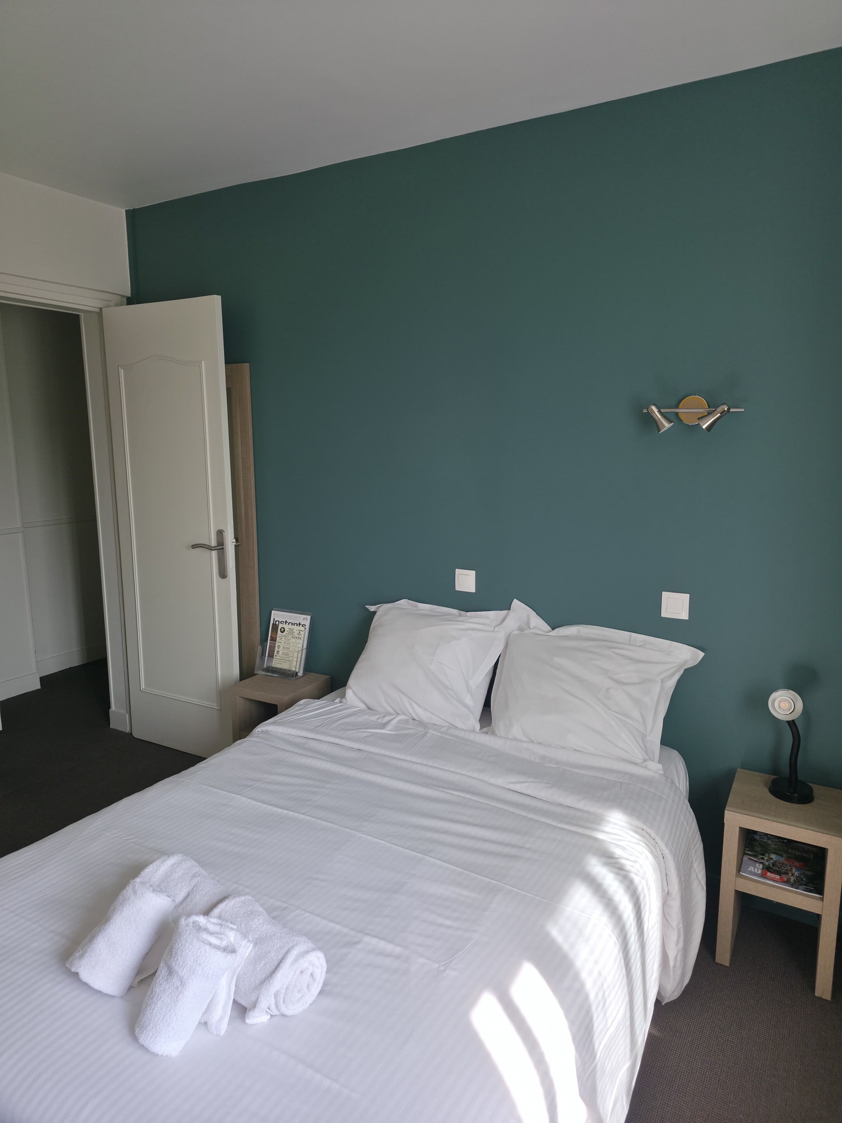 Quarto de hotel com cama, almofadas, mesa de cabeceira, relógio na parede e uma porta.