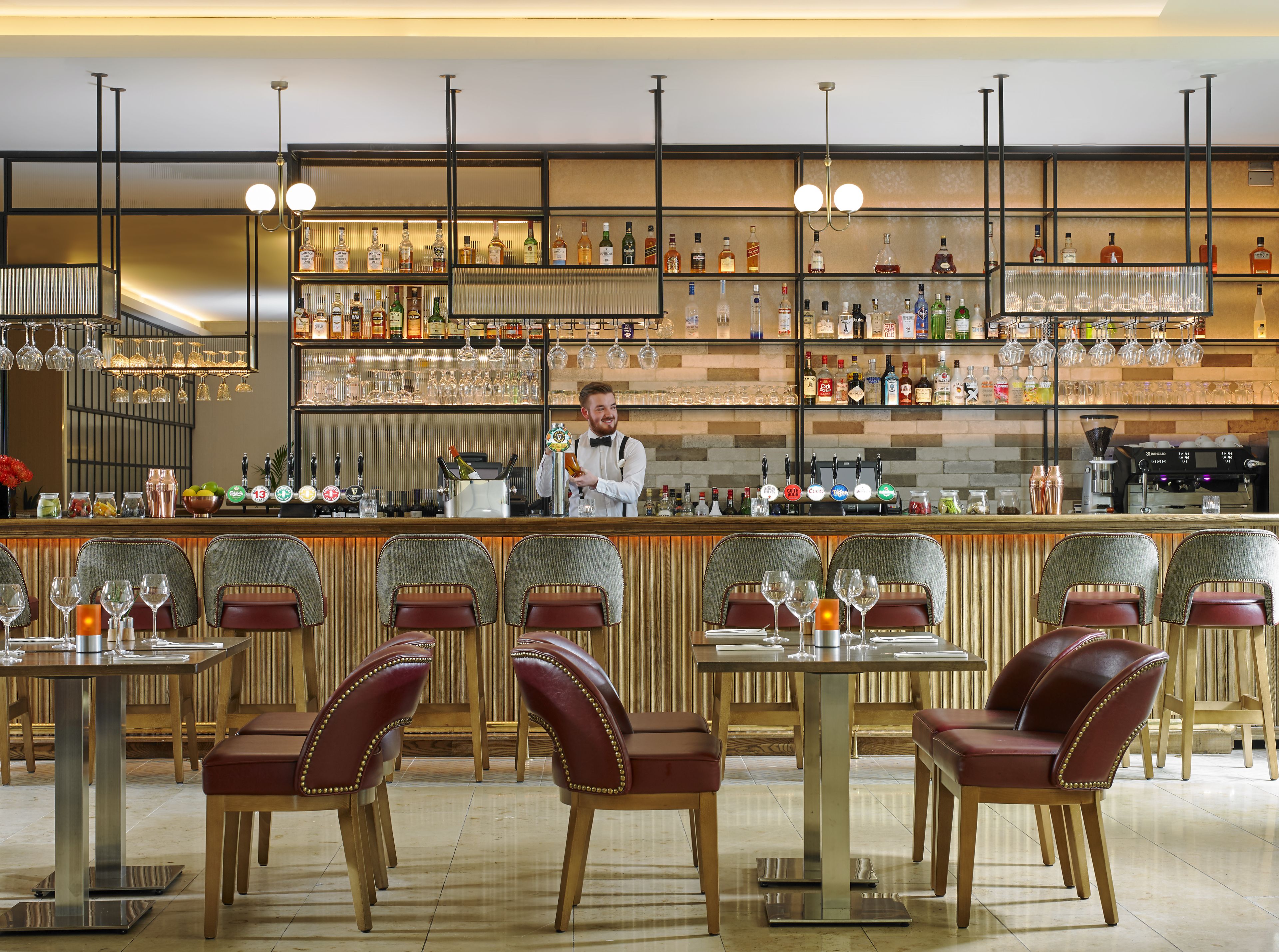 Uma área de bar com um bartender, várias prateleiras de garrafas de bebidas e banquetas.