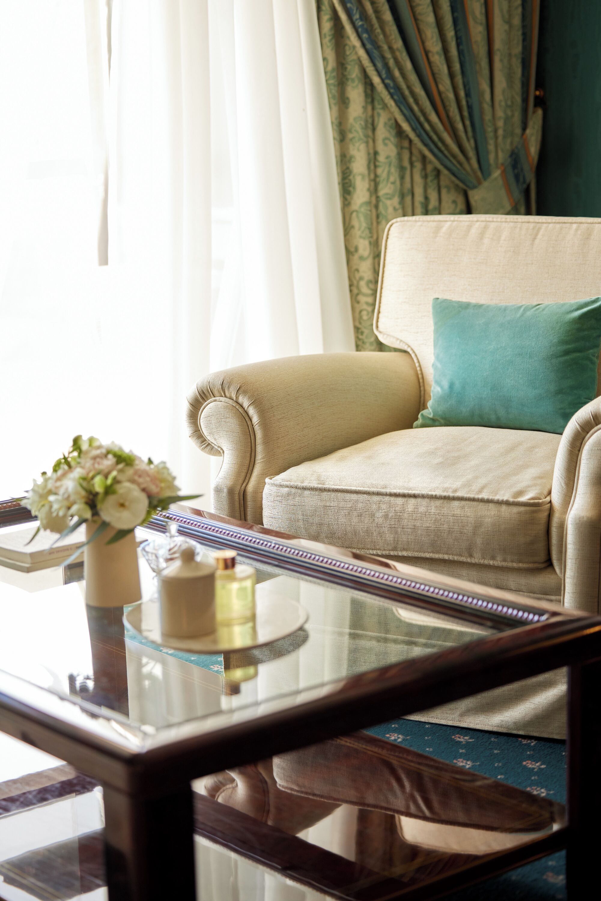 Uma sala de estar com poltrona bege, almofada azul-esverdeada, um vaso com flores e uma mesa de centro de vidro.