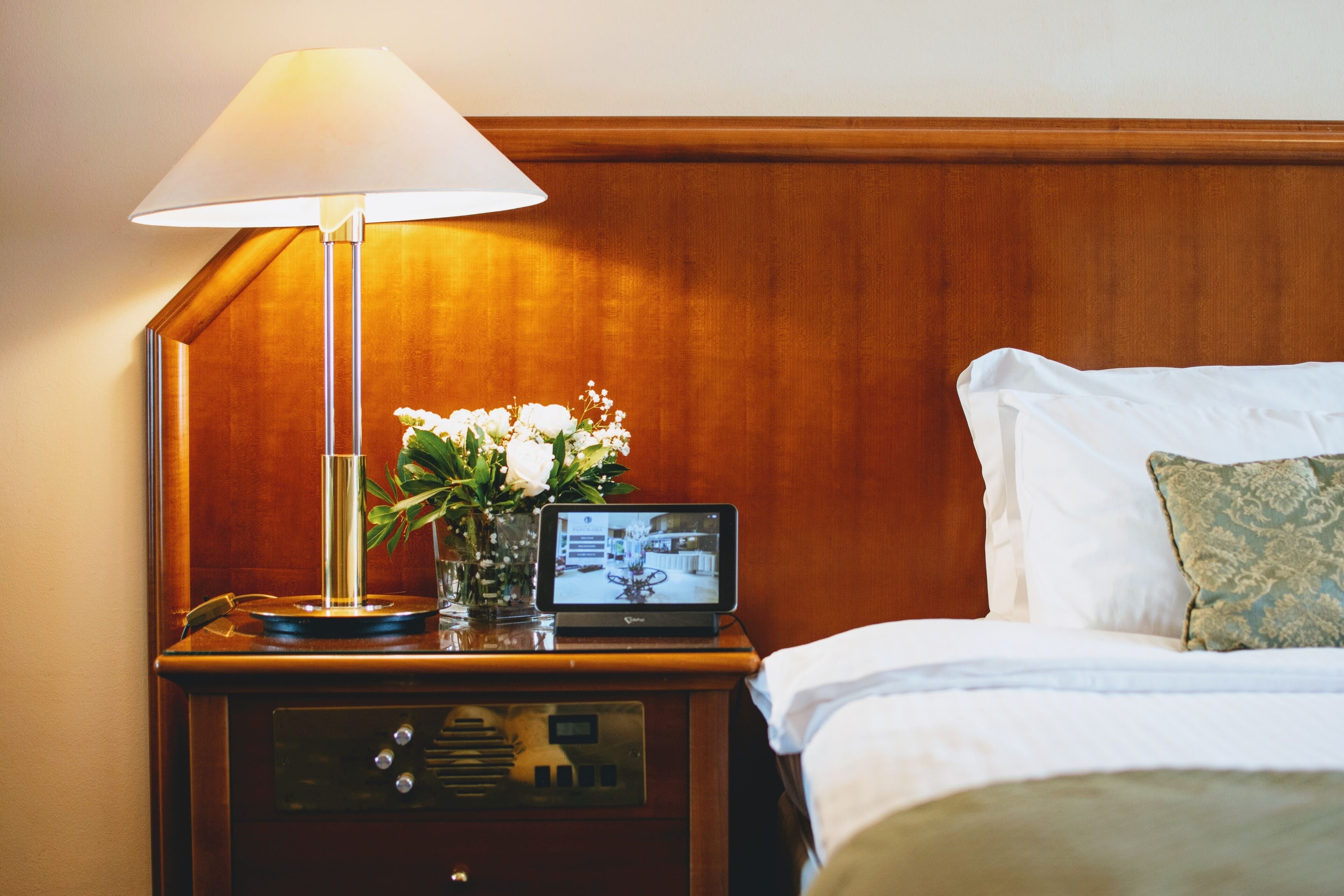 Quarto de hotel com cabeceira de madeira, mesa de cabeceira com luminária, uma TV pequena e um vaso com flores.