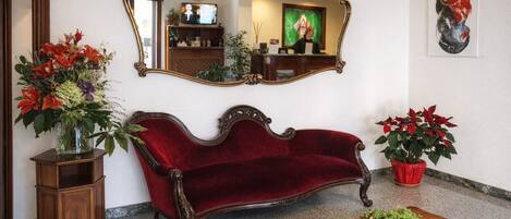Um hall com um sofá de veludo vermelho, uma mesa de centro de madeira, um espelho grande e ornamentado e duas plantas em vasos.