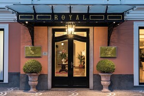 Entrada de hotel com uma porta preta, duas plantas em vasos e uma placa com a inscrição 'ROYAL'.