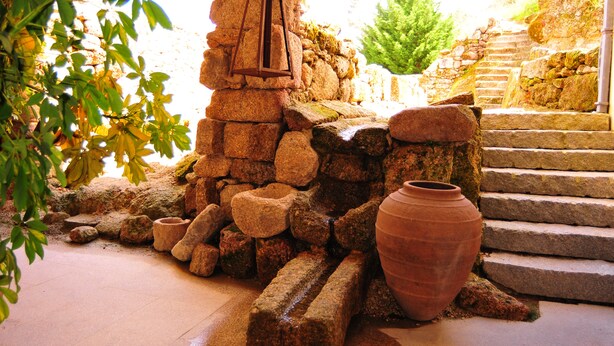 Escadaria de pedra que leva a uma parede de pedra, com um candeeiro e um grande vaso de terracota.