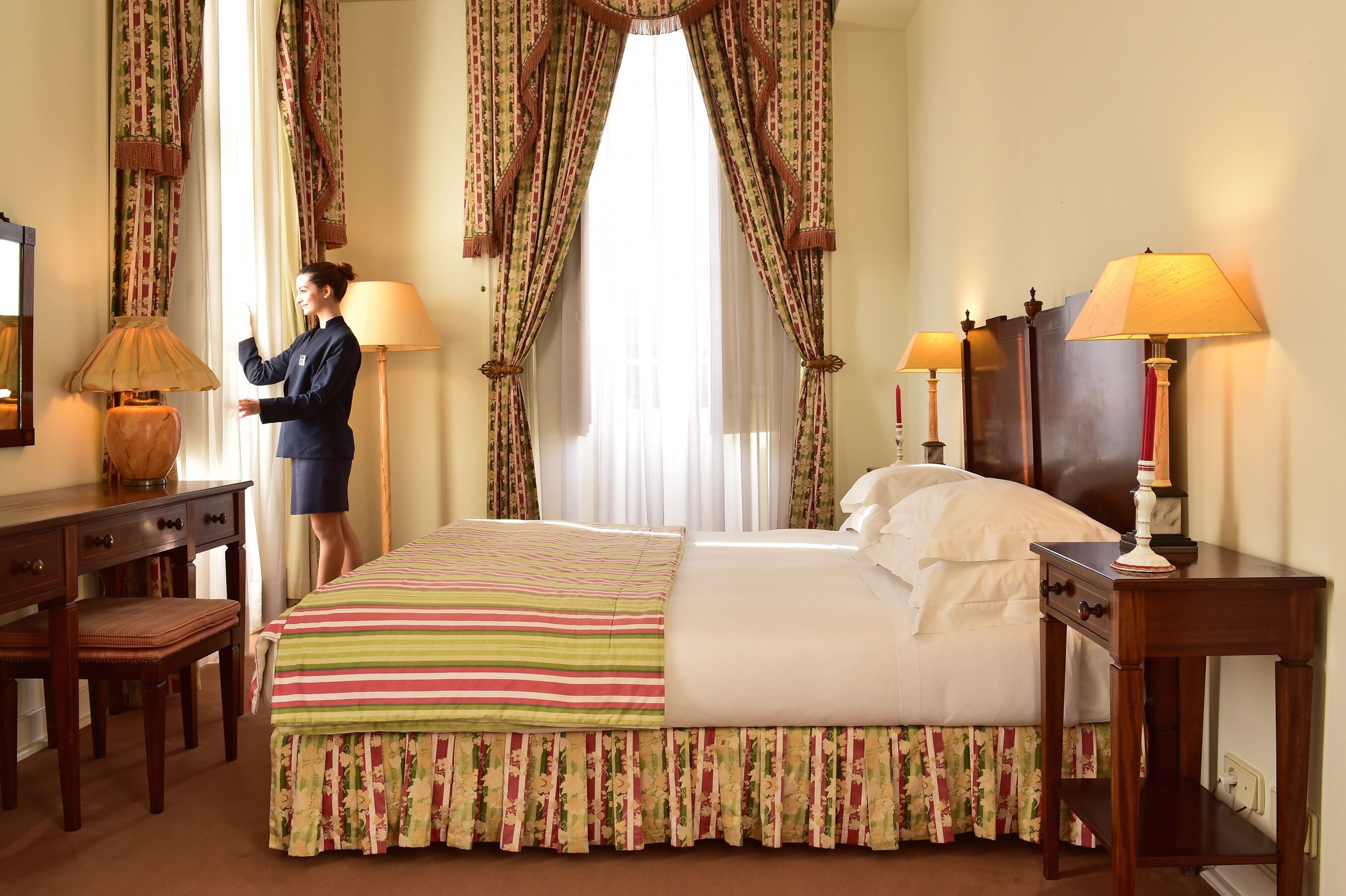 Quarto de hotel com cama, duas mesas de cabeceira, escrivaninha, cadeira e uma janela grande com cortinas.