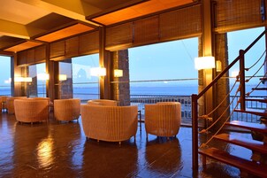 Amplo espaço interno com cadeiras e mesas de vime, grandes janelas com vista para o mar e uma escada de madeira.