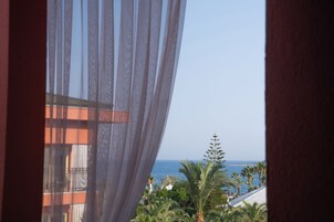 Vista através de cortinas finas, mostrando uma varanda com grades, um edifício com fachada vermelha e uma praia bordeada por palmeiras.