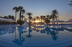 Piscina com espreguiçadeiras e palmeiras, refletindo o pôr do sol.