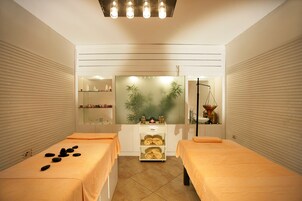 Sala de spa com duas macas de massagem, uma prateleira com toalhas e um espelho.