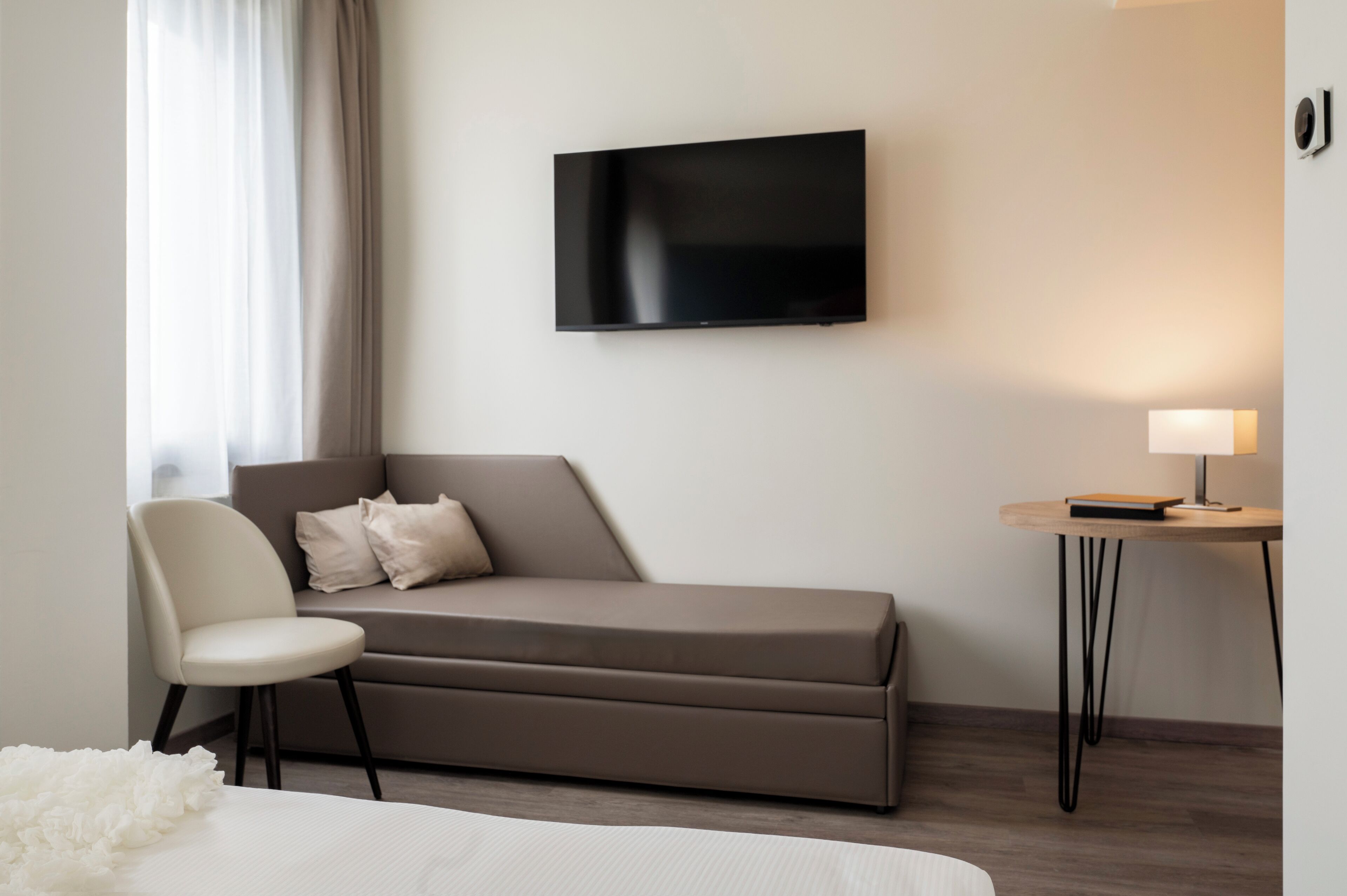 Quarto de hotel moderno com televisão de tela plana na parede, um sofá, uma cadeira, uma mesa pequena com abajur e uma cama com roupa de cama branca.