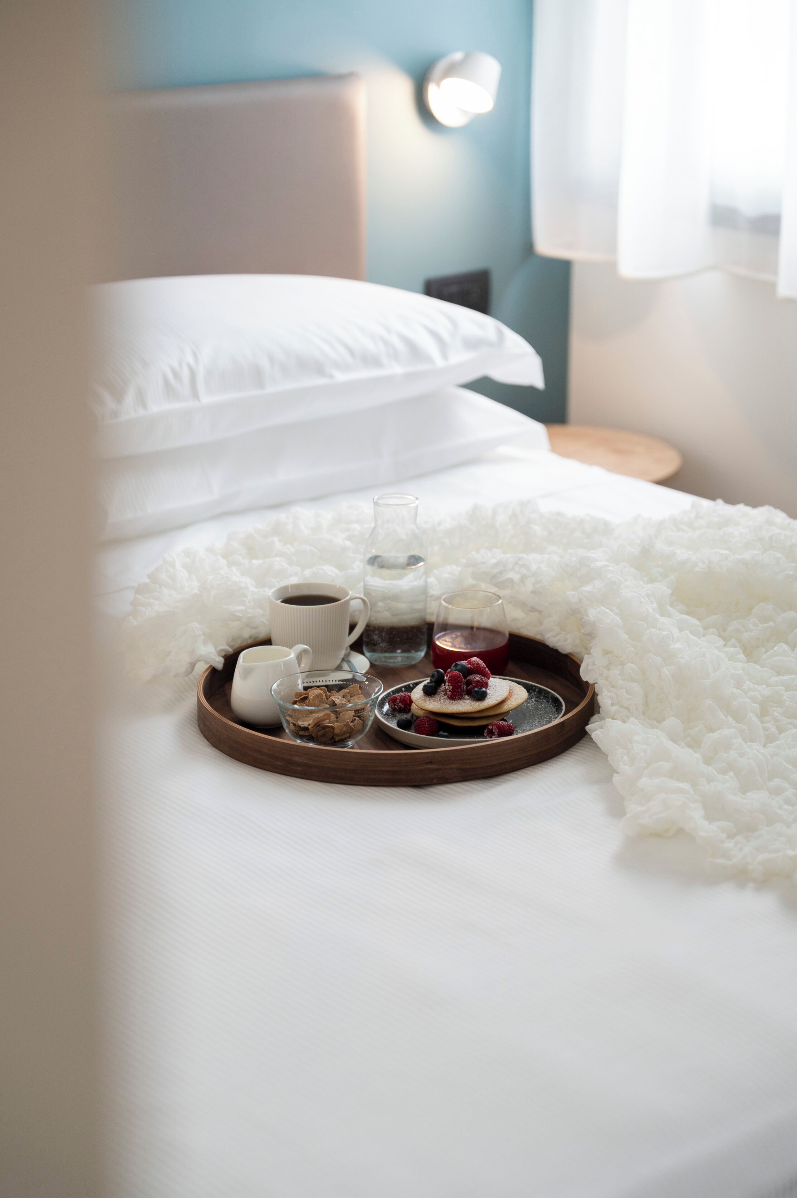 Uma bandeja de café da manhã bem arrumada sobre a cama, com lençol e travesseiro brancos, e uma cabeceira.