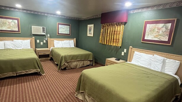 Quarto de hotel com três camas de solteiro, uma pequena mesa de cabeceira com abajur e uma janela grande com cortinas.